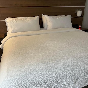 Uma cama bem arrumada com roupa de cama branca, cabeceira de madeira e mesa de cabeceira com relógio e dispensador de lenços.