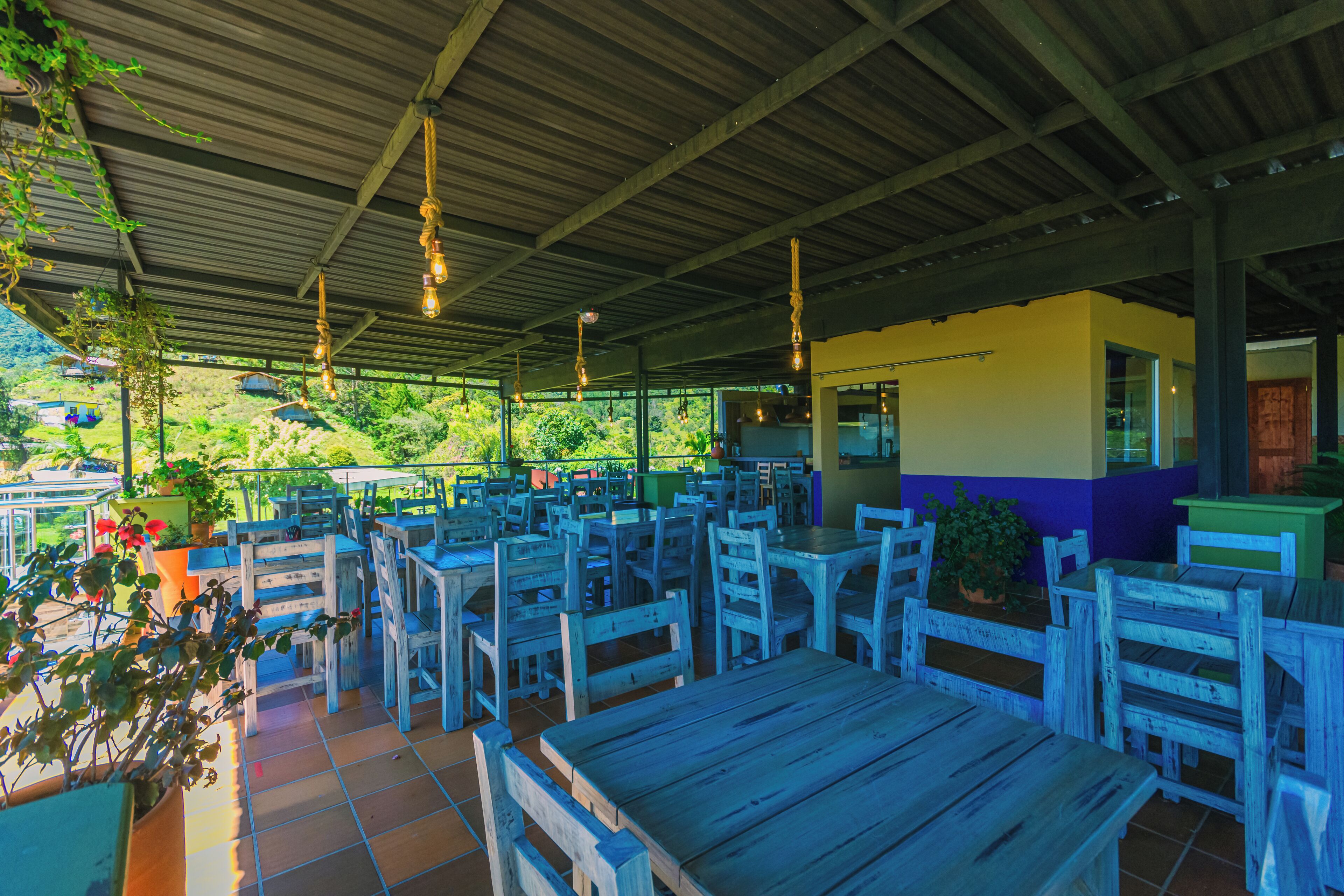 Um restaurante ao ar livre com mesas e cadeiras de madeira, luzes pendentes e plantas em vasos.