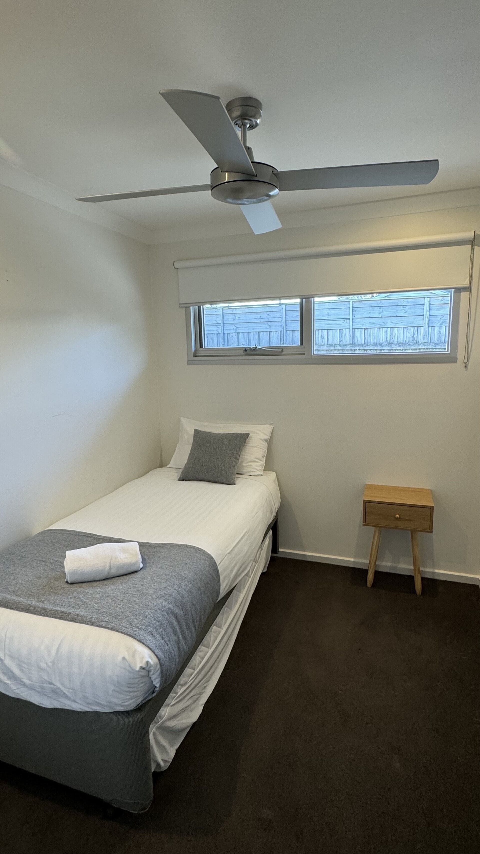 Quarto com cama, ventilador de teto, janela e uma pequena mesa de cabeceira de madeira.