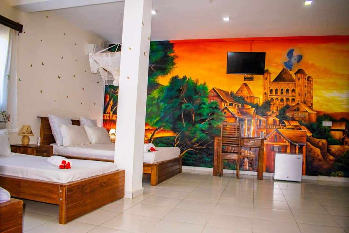 Quarto de hotel com duas camas de madeira, um mural de uma floresta e edifícios, uma TV de tela plana e um frigorífico branco.