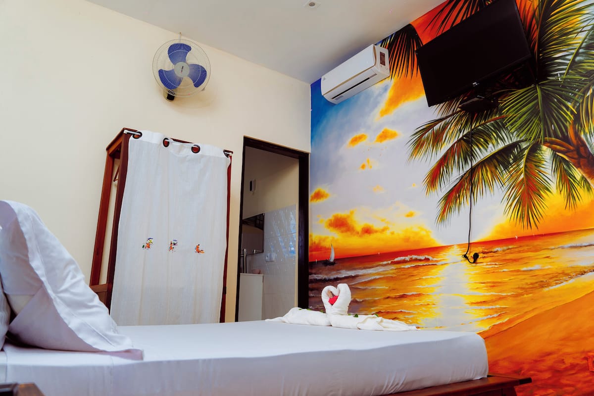 Quarto de hotel com uma cama, um ventilador, um aparelho de ar condicionado, um guarda-roupa e um mural na parede com uma cena de praia.