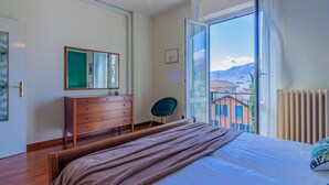 Quarto com cama, cômoda, cadeira e uma janela grande com vista para prédios e montanhas.