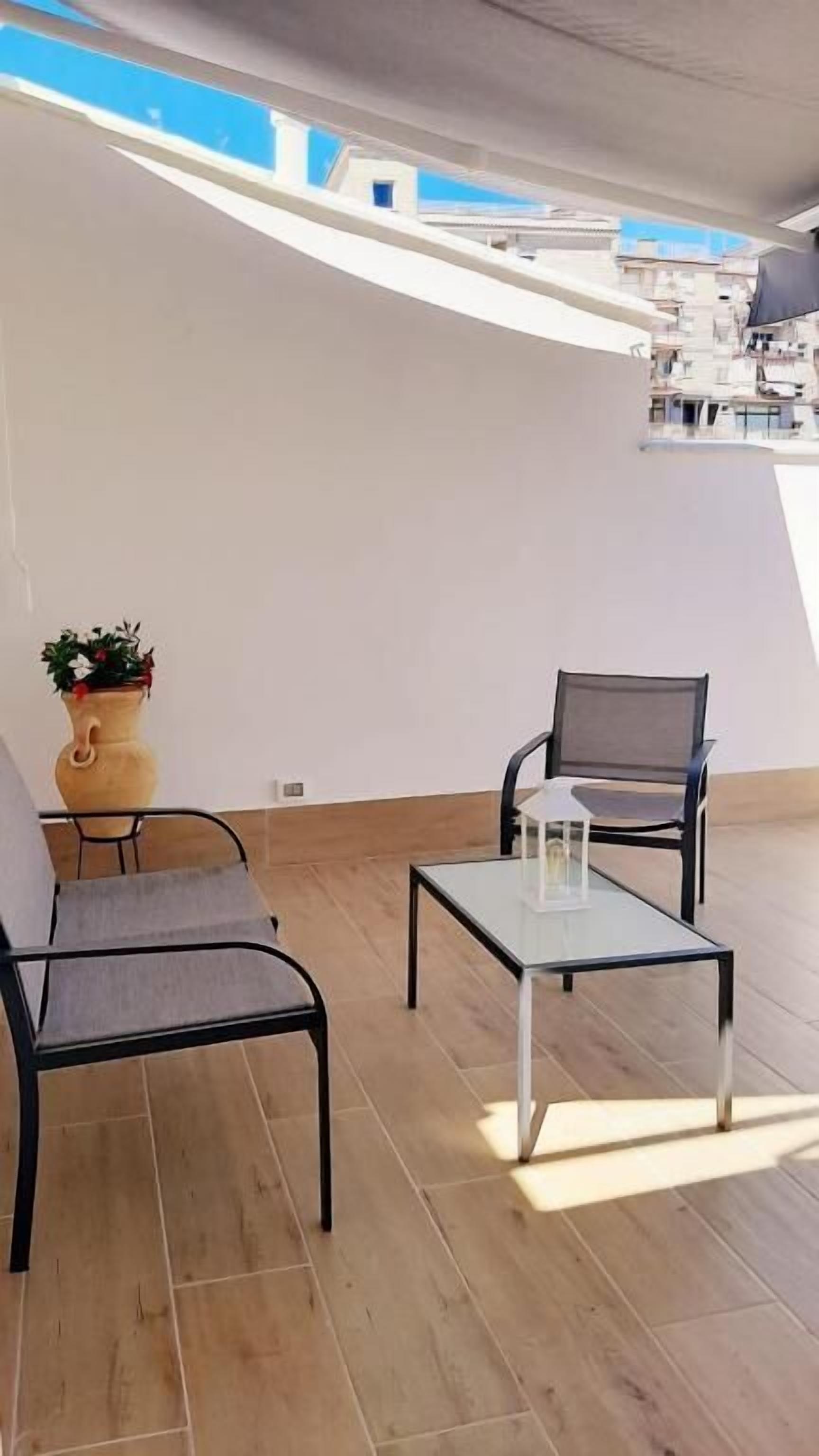 Uma área de estar moderna ao ar livre, com duas cadeiras, uma mesa e um vaso grande com flores, além de vista para edifícios ao fundo.