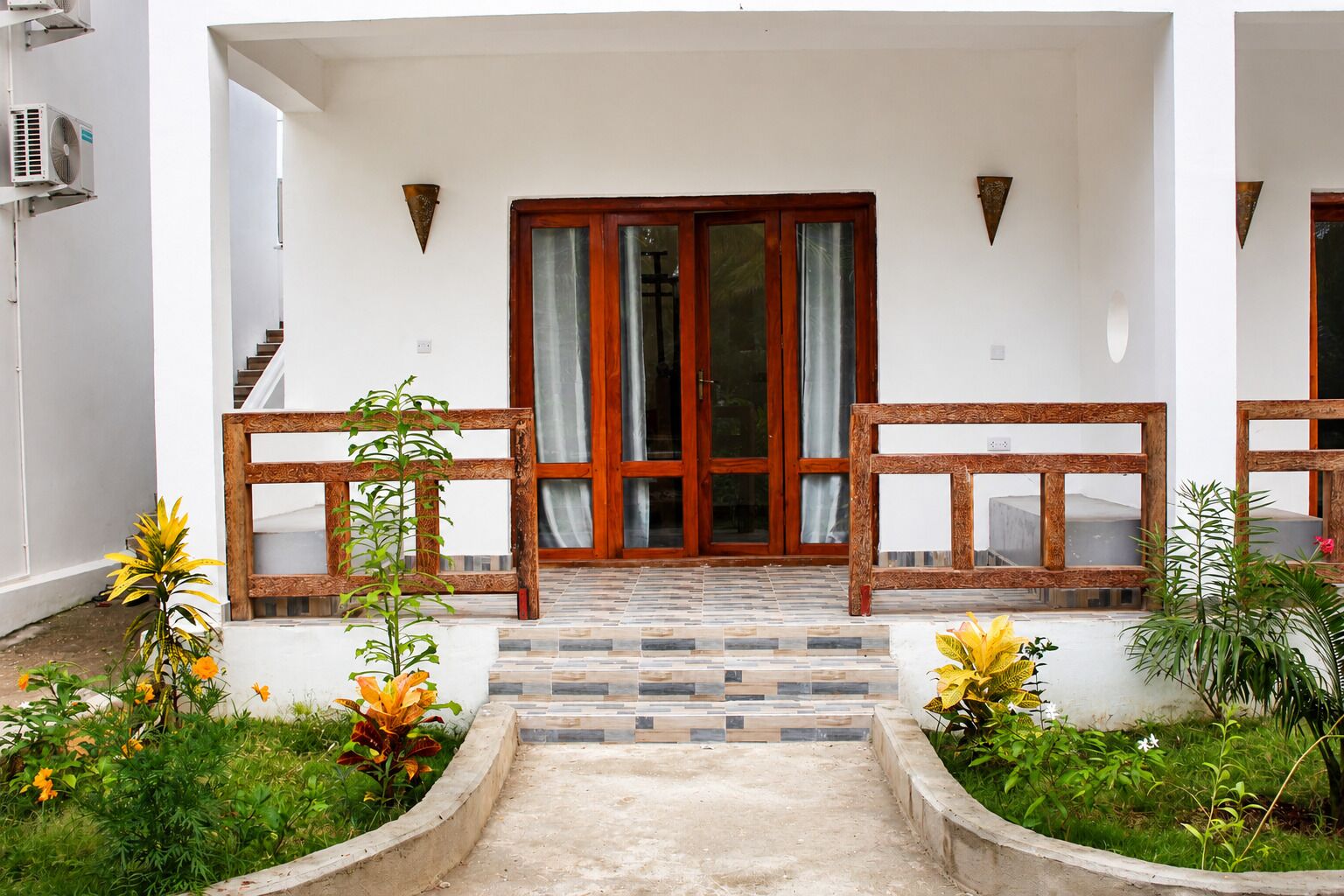 Um edifício com porta de madeira, escadaria que dá para uma varanda e um jardim com diversas plantas.