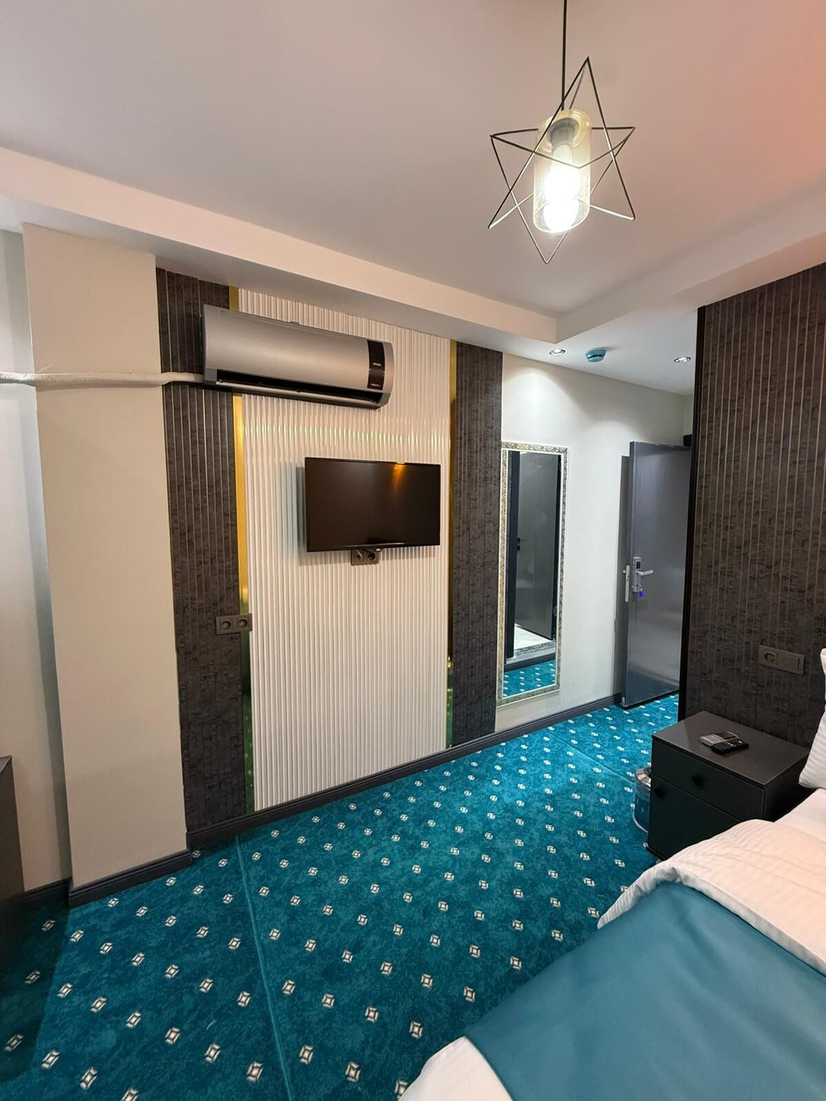 Quarto de hotel com cama, televisão fixada na parede, espelho, aparelho de ar condicionado e luminária em formato de estrela.