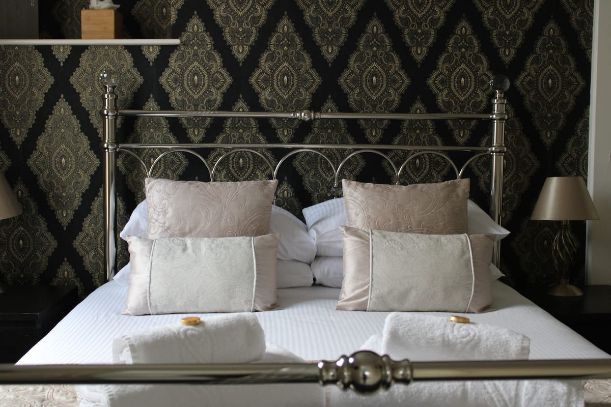 Cama bem arrumada com estrutura de metal, várias almofadas e duas luminárias de cabeceira.