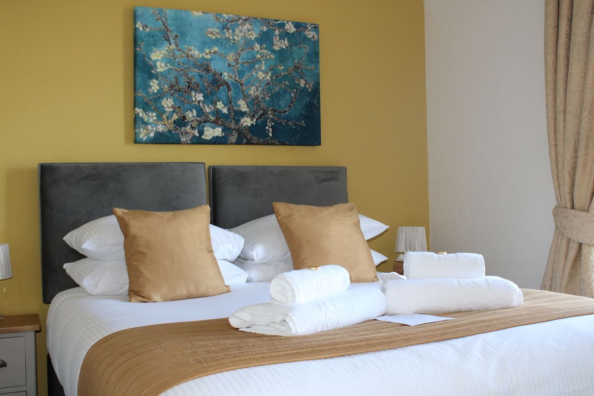 Cama bem arrumada com lençóis brancos e duas almofadas castanhas, um quadro floral azul na parede e uma cortina bege na janela.