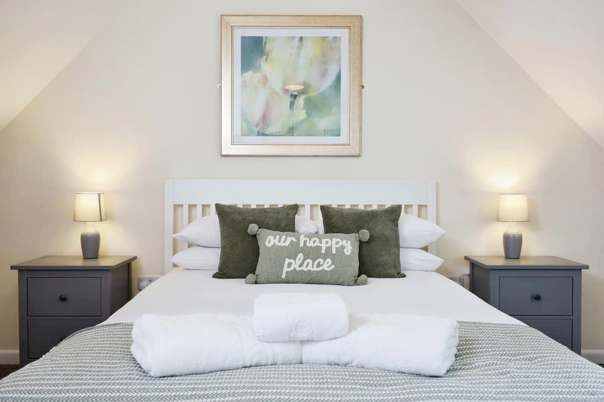 Cama bem arrumada com um travesseiro que diz "nosso lugar feliz", duas mesas de cabeceira com abajures e um quadro abstrato emoldurado acima da cama.
