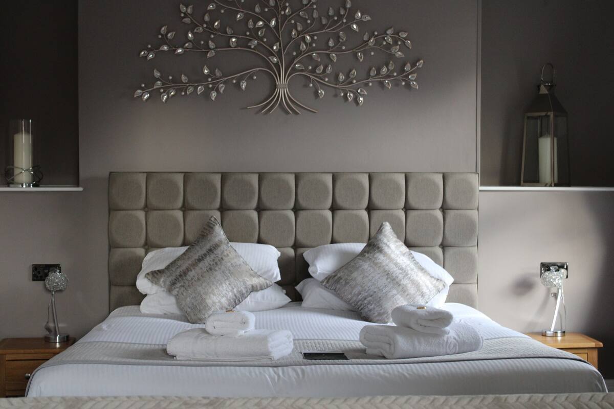 Cama bem arrumada com cabeceira estofada, duas almofadas decorativas e um conjunto de toalhas dobradas.