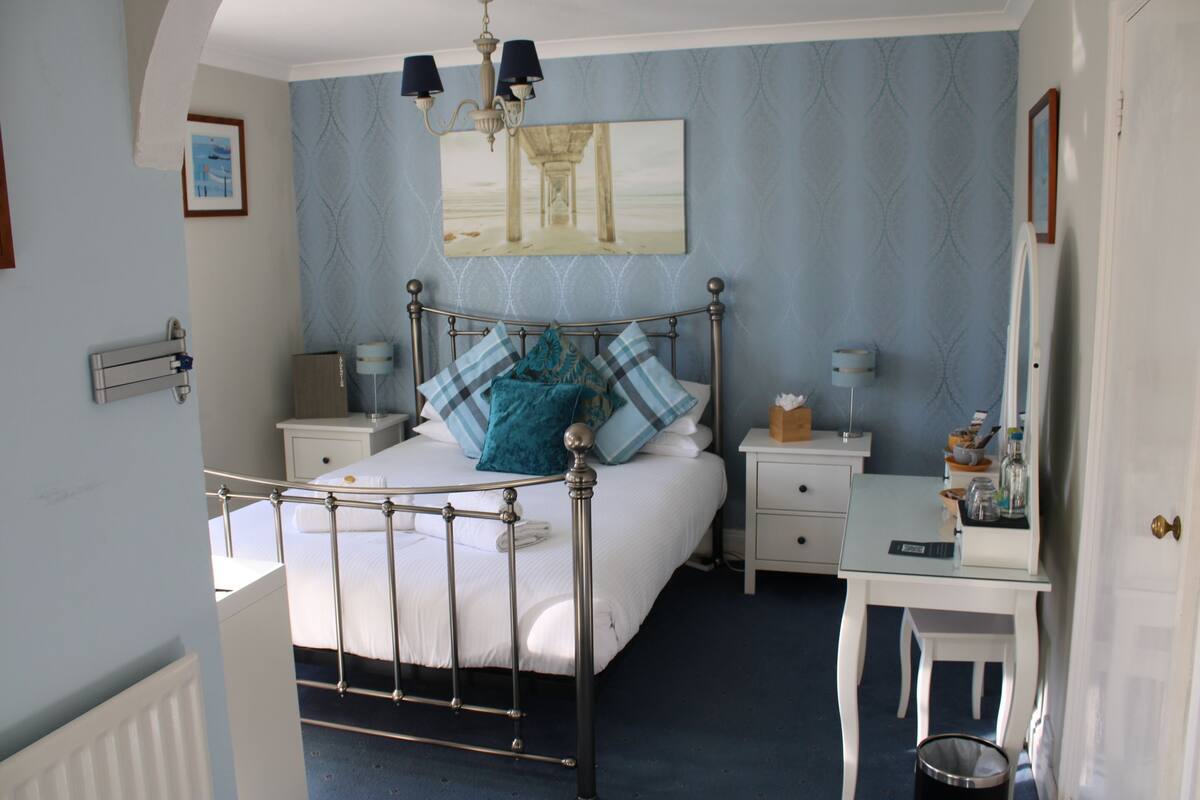 Quarto com estrutura de cama de metal, mesa de cabeceira com abajur, escrivaninha com cadeira e quadro na parede.
