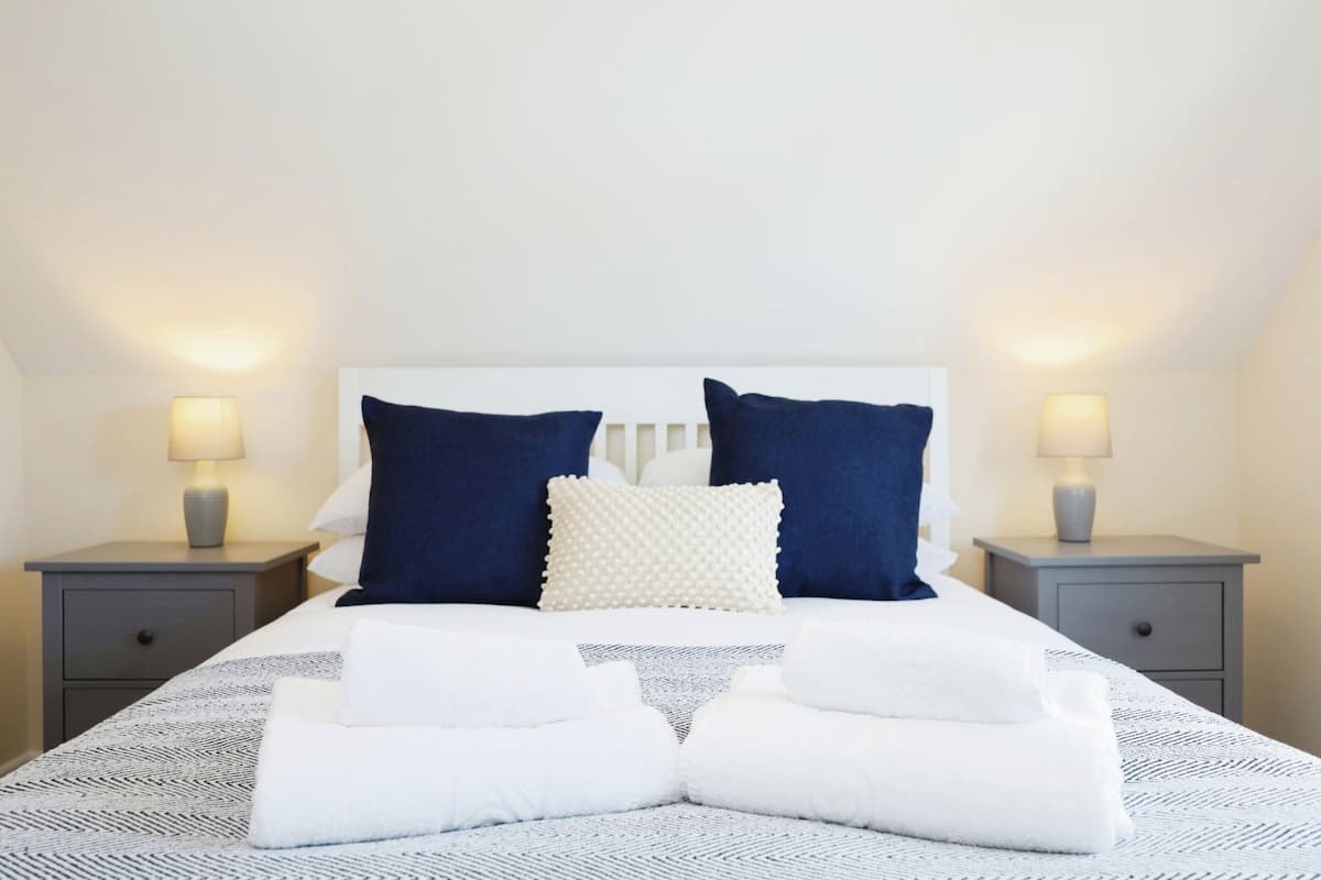 Cama bem arrumada com almofadas brancas e azul marinho, cabeceira branca e duas mesas de cabeceira com abajures.