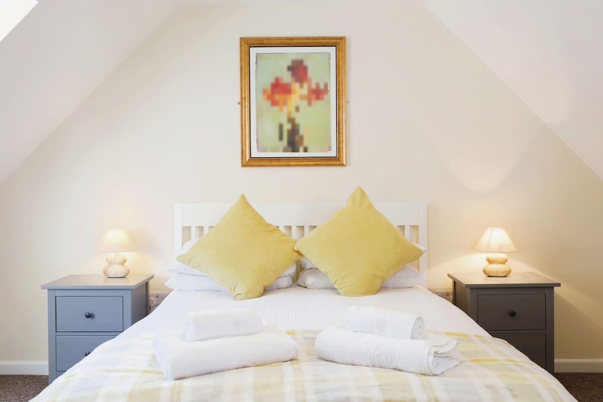 Cama bem arrumada com lençóis brancos e duas almofadas amarelas, com duas mesas de cabeceira com abajures, em um quarto com teto inclinado e uma obra de arte emoldurada acima da cama.