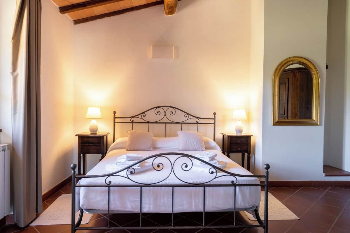 Uma cama bem arrumada com estrutura de metal, duas mesas de cabeceira com luminárias, um espelho na parede e piso de azulejos.
