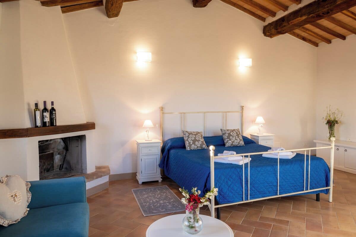 Quarto com cama, mesas de cabeceira, um sofá, lareira e um vaso com flores.