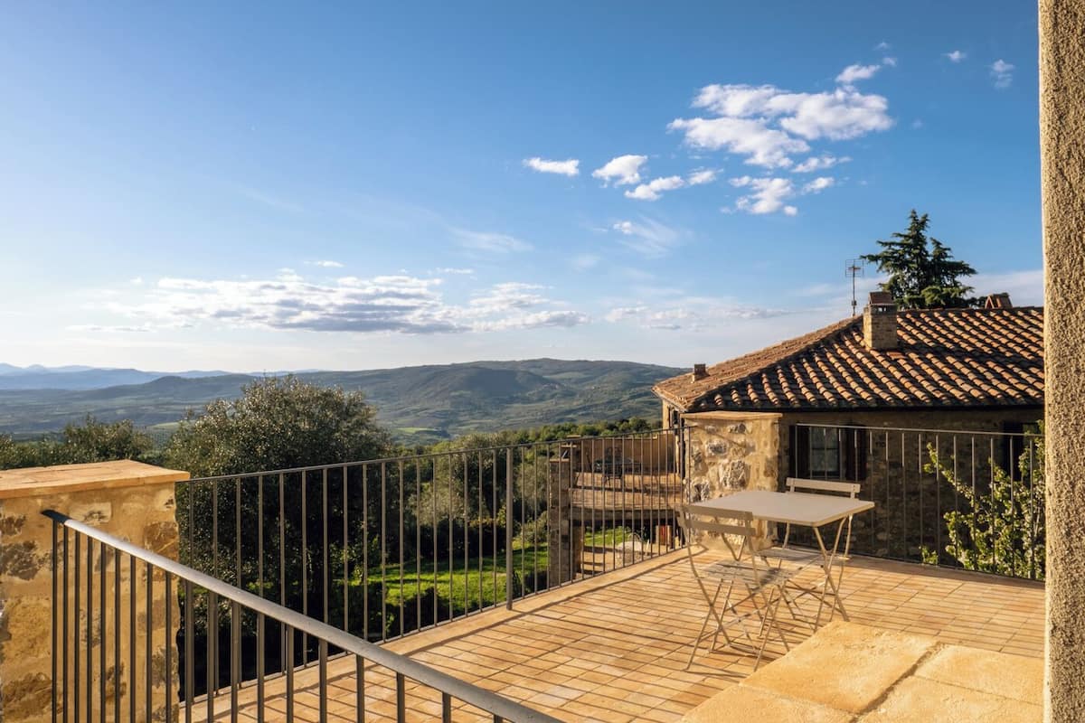 Um terraço com vista para colinas ondulantes, um edifício de pedra com telhado de telhas e um guarda-corpo de metal.