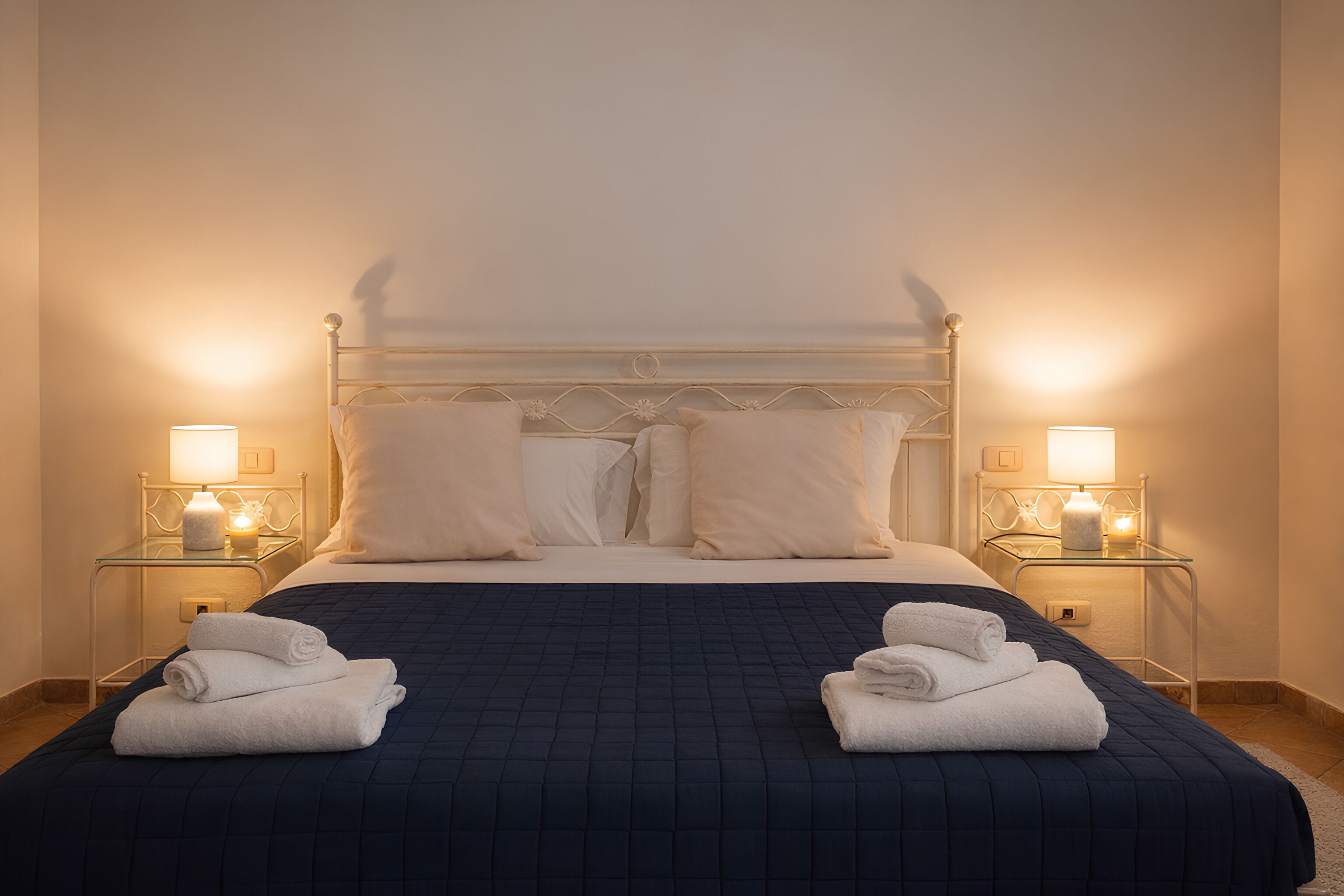 Cama bem arrumada com colcha azul escura, duas mesas de cabeceira com luminárias e toalhas dobradas na cama.