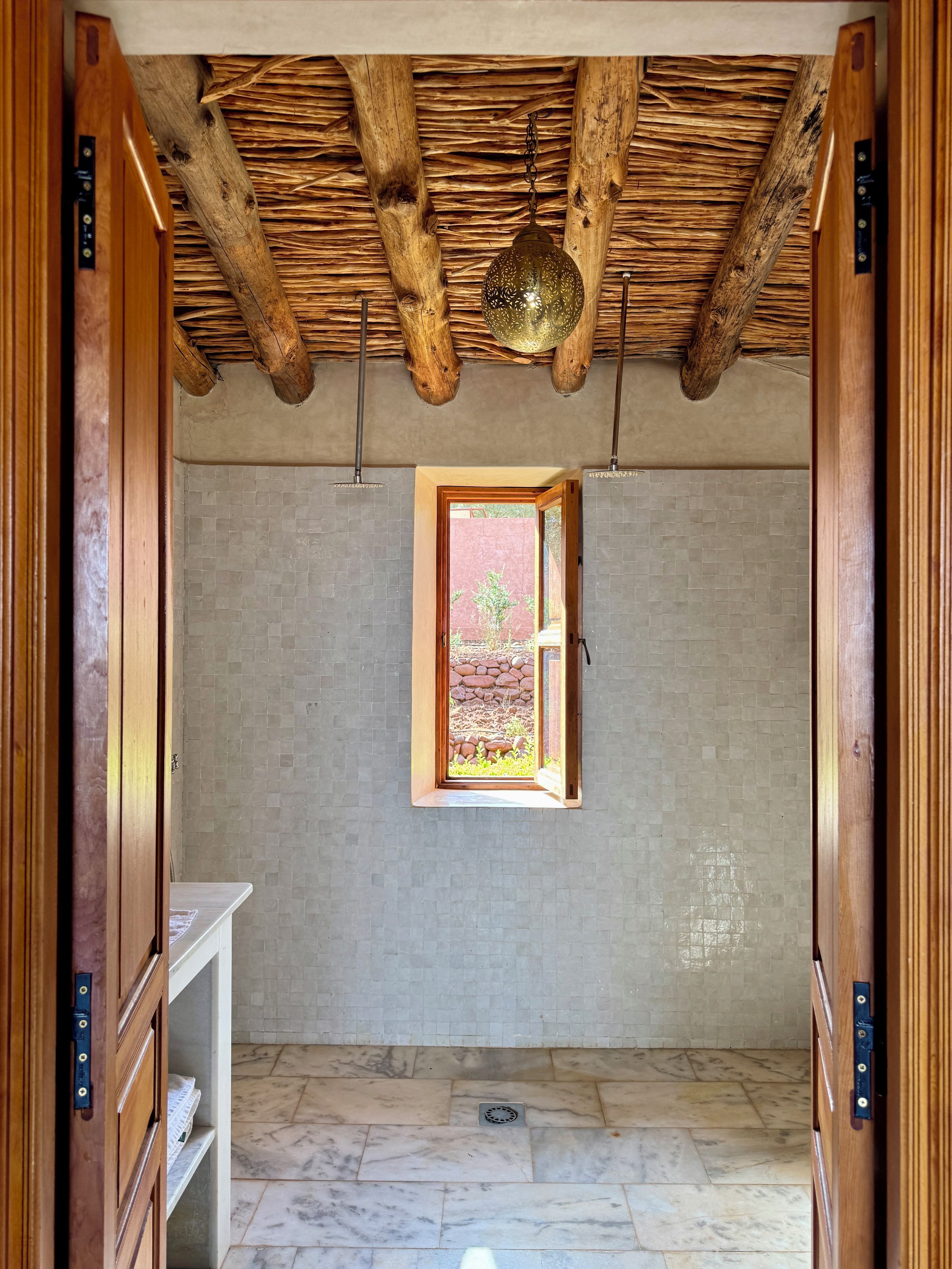 Quarto com teto de madeira rústica, pequena janela com vista para o verde e piso de azulejos.