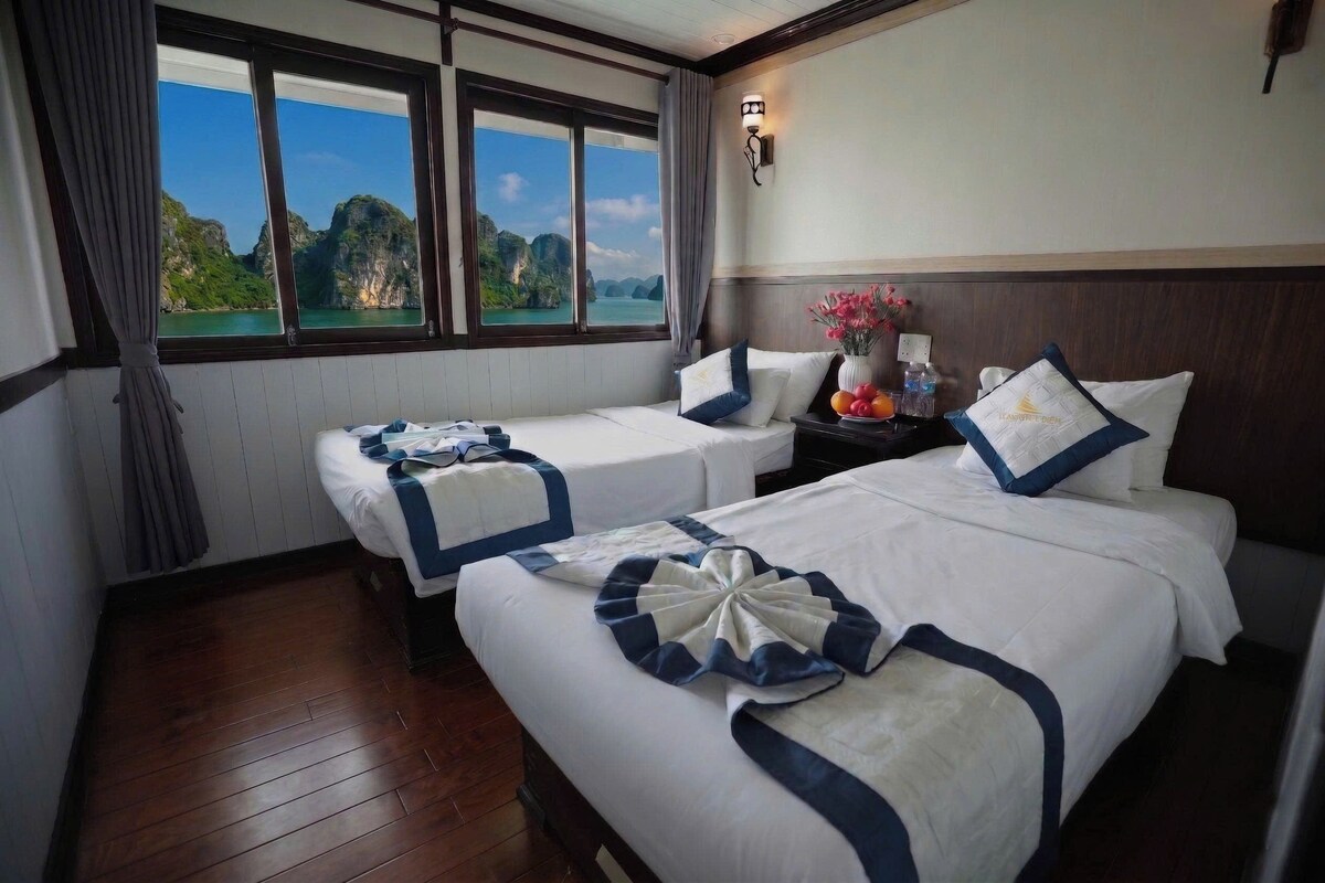 Quarto de hotel com duas camas, mesa de cabeceira, vaso com flores e vista para a paisagem costeira através de grandes janelas.