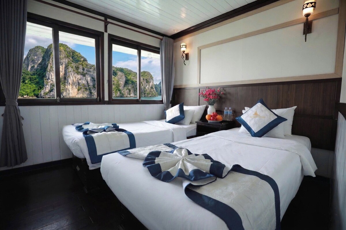 Quarto de hotel com duas camas, uma mesinha de cabeceira, um vaso com flores e vista para uma paisagem montanhosa através de janelas amplas.