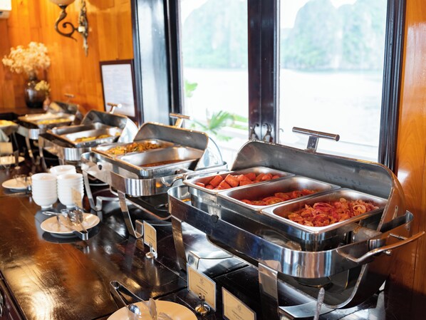 Buffet montado com diversas estações de comida, área de bar e vista para o exterior através de grandes janelas.