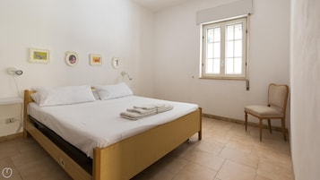 Quarto com cama de madeira, roupa de cama branca, uma cadeira e uma janela.