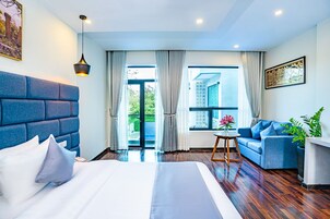 Quarto de hotel moderno com uma cama grande, um sofá azul, uma mesa pequena com flores e vista para o exterior através de amplas janelas.