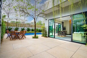 Um pátio externo com piscina, cadeiras de madeira e porta de vidro que dá para uma varanda.