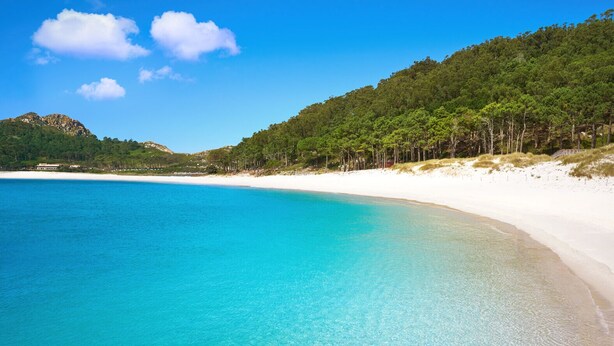 Uma praia com águas azul-turquesa cristalinas, areia branca e um morro coberto por vegetação.