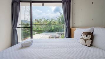 Quarto de hotel com uma cama grande, uma varanda com vista para vegetação e uma linha de energia, e um travesseiro macio em formato de coruja.