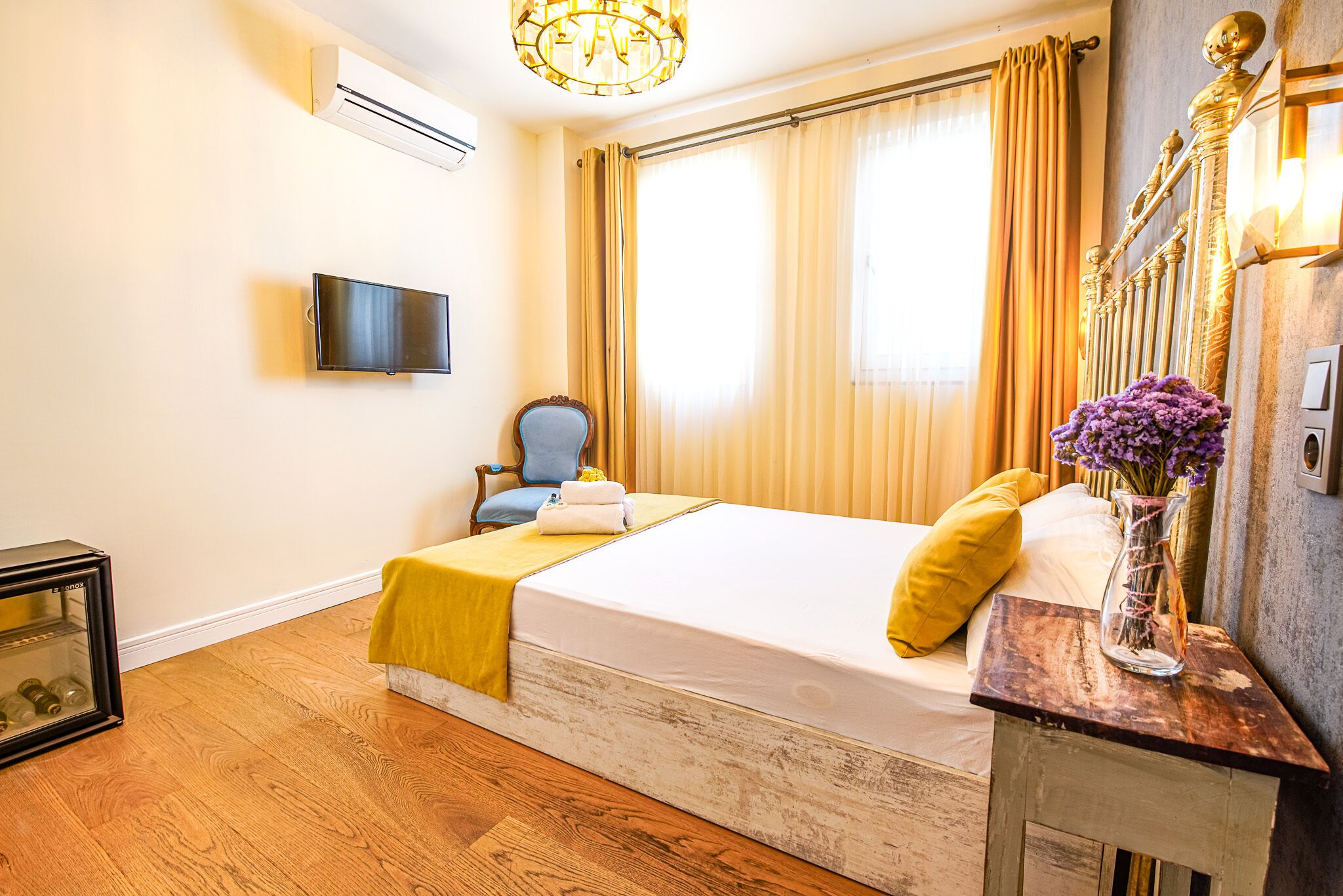 Quarto de hotel com cama, televisão, cadeira e mesa de cabeceira com um vaso.