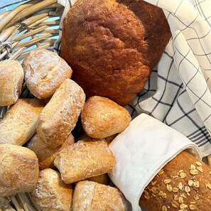 Uma cesta com diversos pães e doces, incluindo um pão grande e vários pãezinhos, sobre um tecido xadrez.