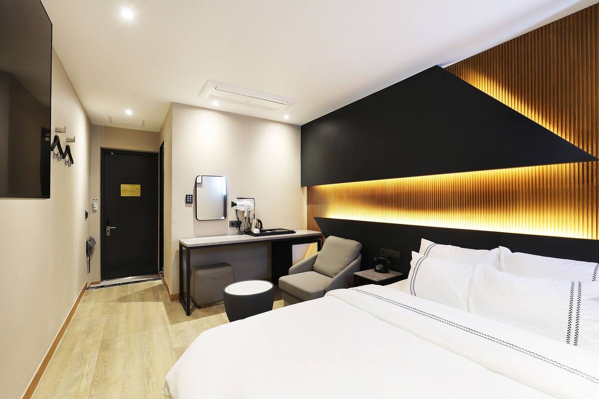 Quarto de hotel moderno com uma cama grande, uma escrivaninha com cadeira, uma mesinha e um espelho fixado na parede.