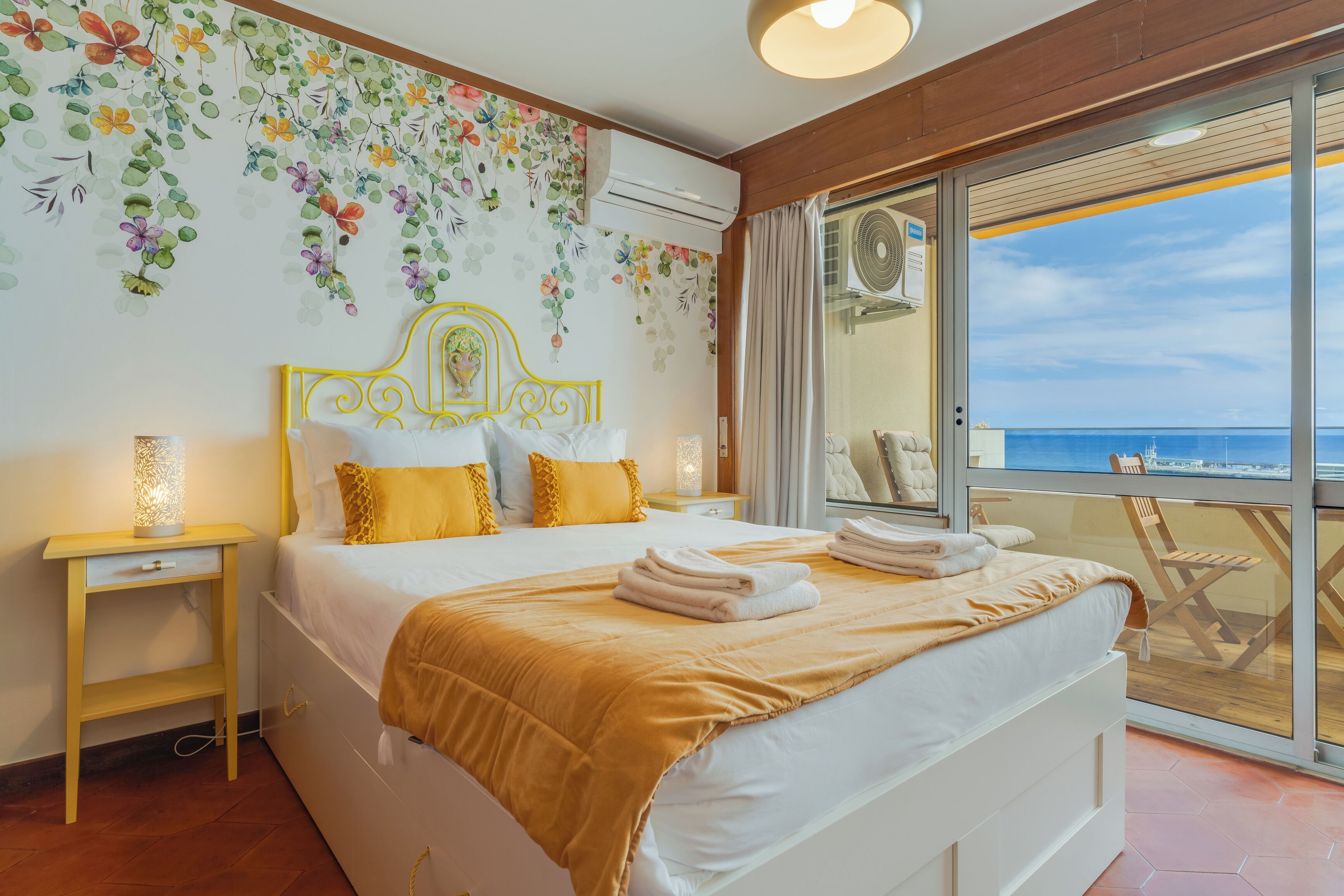 Quarto de hotel com cama, mesa de cabeceira, uma grande janela e vista para o mar, e um mural decorativo na parede.