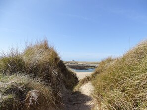 Um caminho arenoso estreito ladeado por grama alta que leva a uma praia com águas calmas e uma construção ao longe.