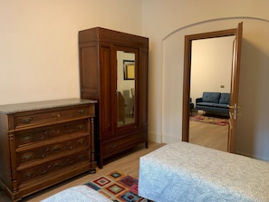 Quarto com cômoda de madeira, cama com colcha estampada e sofá azul ao fundo.
