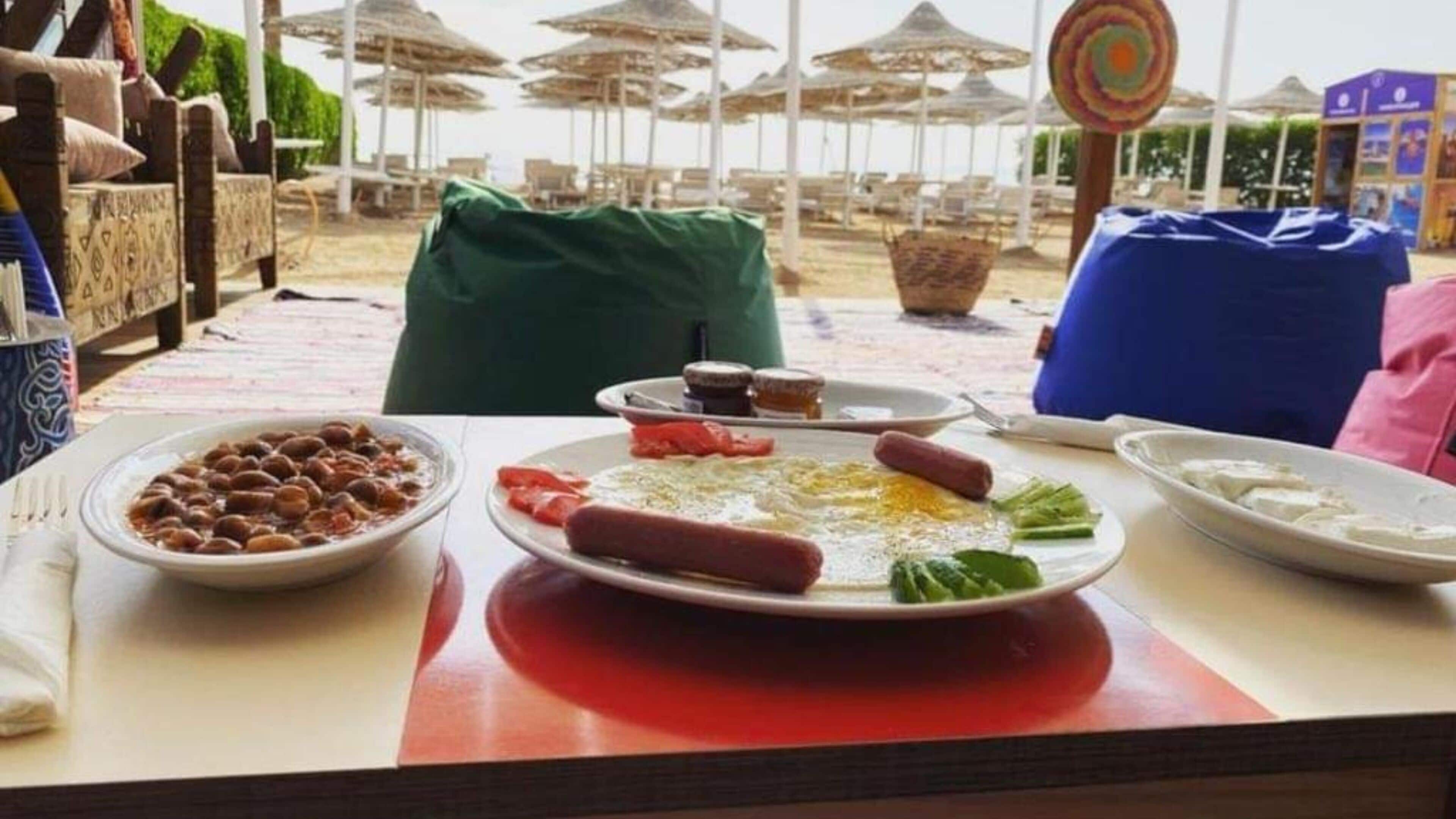 Mesa com itens de café da manhã, incluindo ovos, salsichas e vegetais, em um restaurante à beira-mar, com guarda-sóis e cadeiras de praia ao fundo.