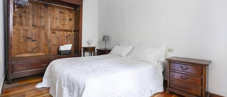 Quarto com um amplo guarda-roupa de madeira, cama com roupa de cama branca, uma pequena mesa de cabeceira com abajur e um criado-mudo de madeira.