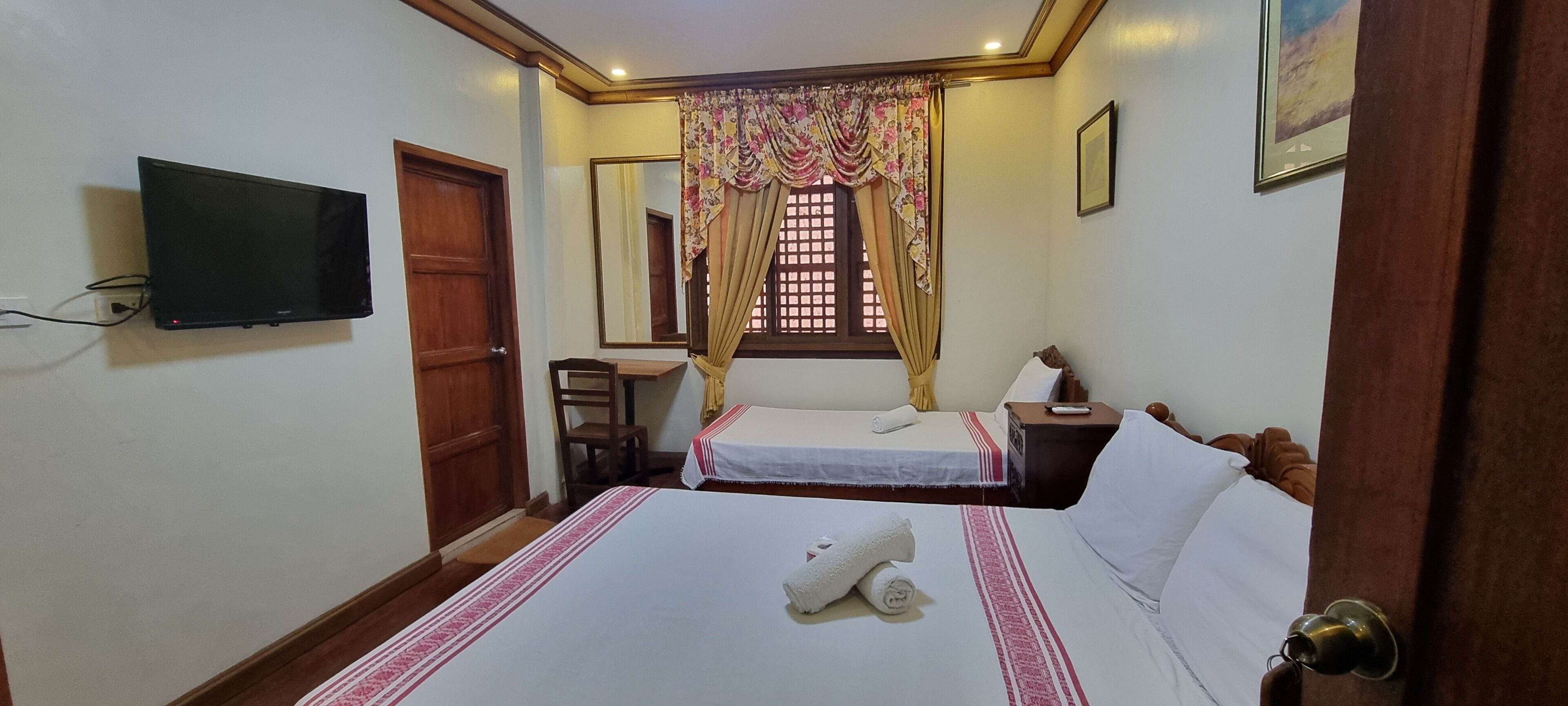 Quarto de hotel com duas camas, televisão na parede, escrivaninha e janela com cortinas.