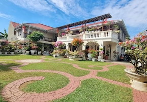 Um edifício de dois andares com telhado de telhas vermelhas, varanda com plantas em vasos e um jardim bem cuidado com caminho de tijolos.