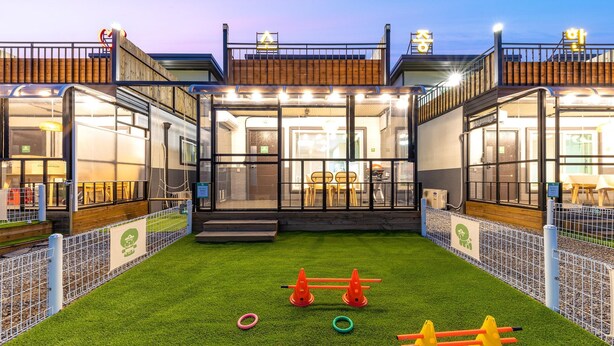 Um edifício moderno com fachada de vidro, um pátio ajardinado com mobiliário de exterior e uma placa indicando um restaurante.
