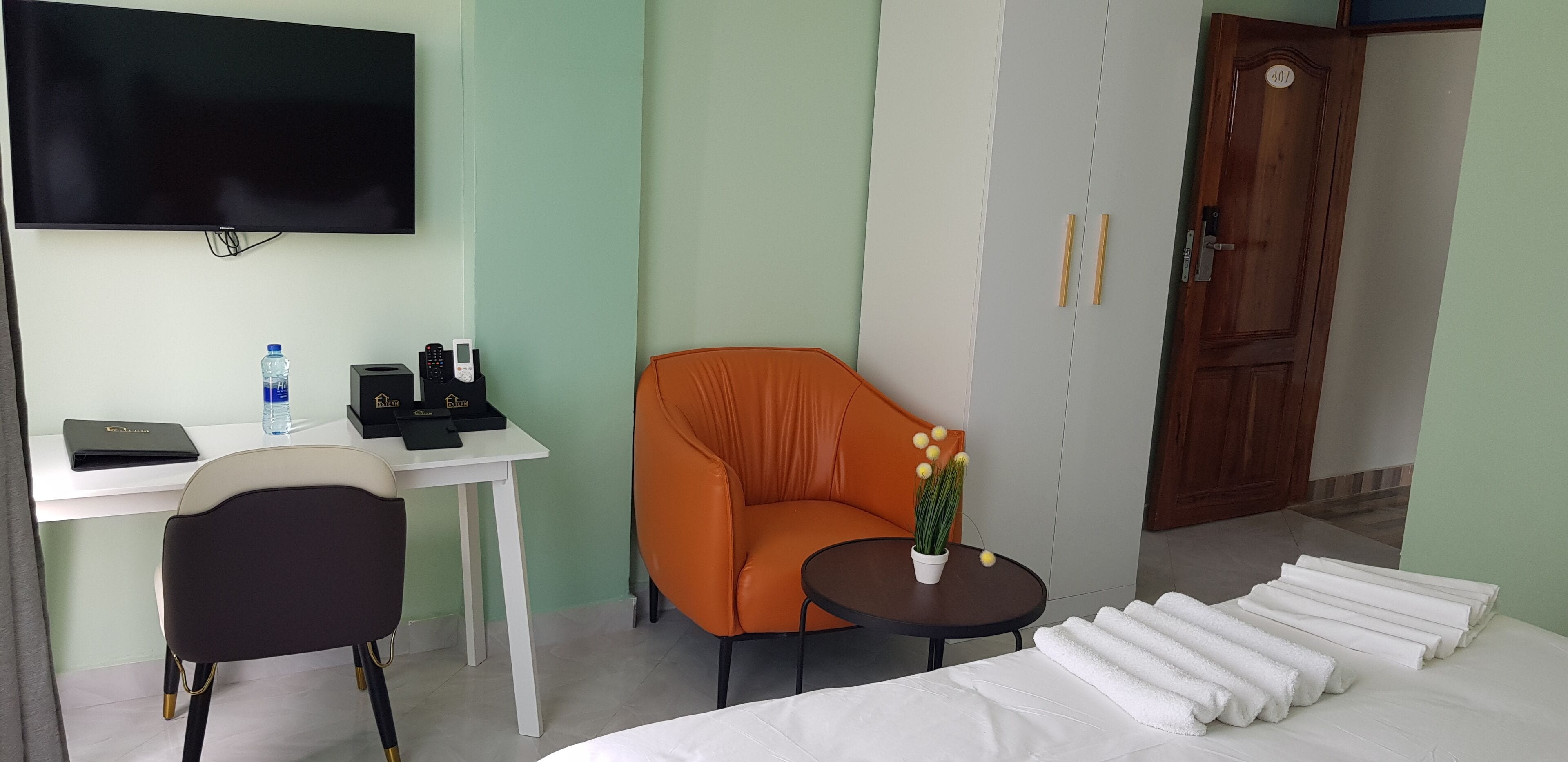 Quarto de hotel com cama, escrivaninha com computador, cadeira, mesinha com planta e porta com número.