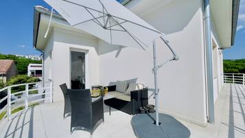 Varanda com parede branca, um sofá, duas cadeiras, uma mesa com bebidas e um grande guarda-sol.