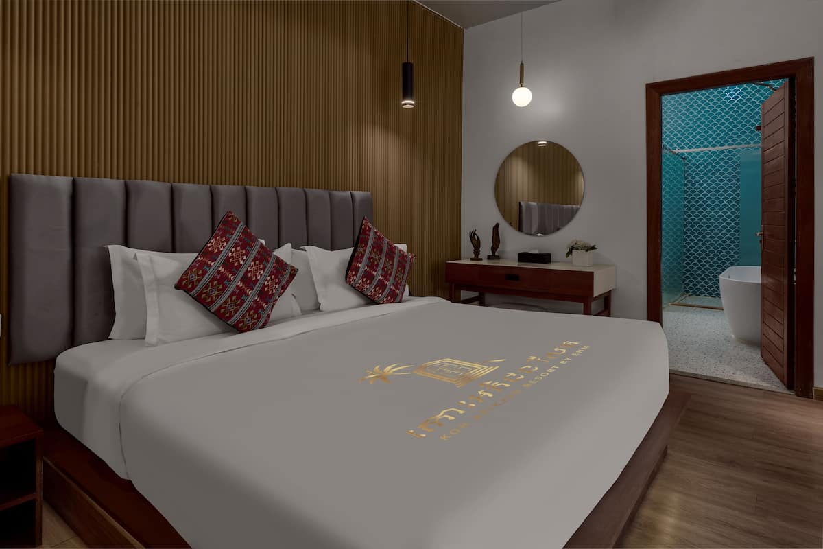 Quarto de hotel moderno com uma cama grande, cabeceira de madeira, mesa de cabeceira com espelho e banheiro visível por uma porta aberta.