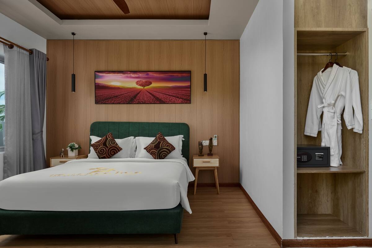 Quarto com uma cama grande, cabeceira de madeira, mesa de cabeceira e um roupão pendurado em um suporte. Há também uma pintura de um pôr do sol sobre um campo.
