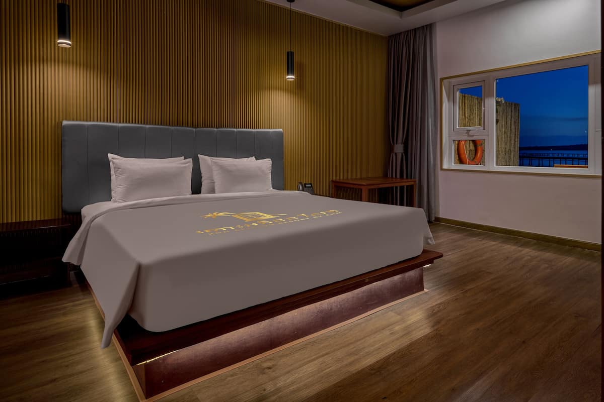 Quarto de hotel com uma cama grande, paredes de painel de madeira, uma mesa pequena e uma janela com vista para o mar.