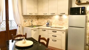 Uma cozinha compacta com armários brancos, micro-ondas, frigorífico e uma mesa de jantar com cadeiras.