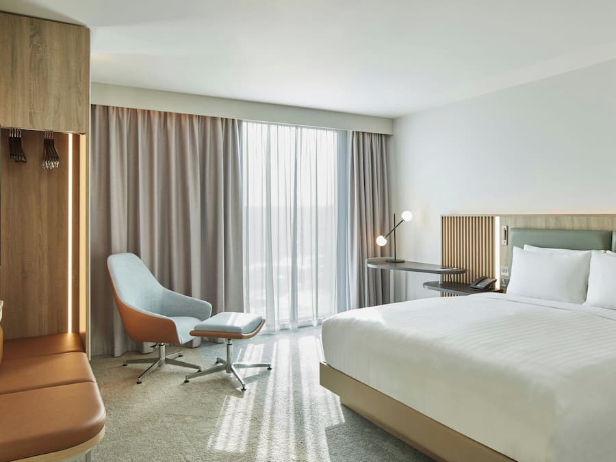 Quarto de hotel com uma cama grande, área de estar com uma poltrona e apoio para os pés, escrivaninha com luminária e televisão na parede.