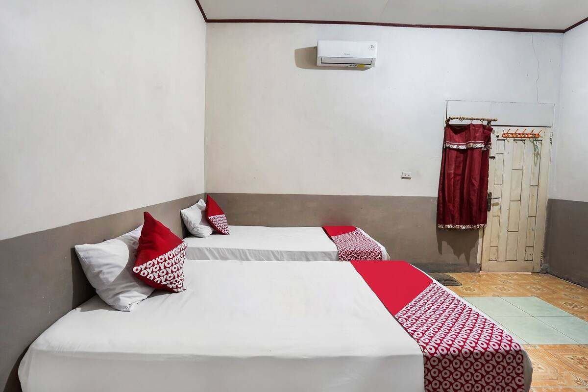 Quarto com duas camas, cada uma com roupa de cama branca e detalhes vermelhos, ar condicionado fixado na parede e uma porta com cortina vermelha.
