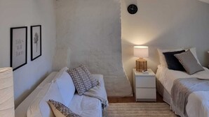 Um quarto aconchegante com teto inclinado, vigas de madeira aparentes, uma cama com edredom branco, uma mesa de cabeceira com abajur e um relógio de parede.
