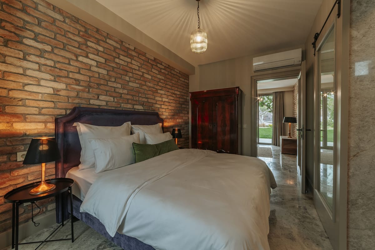 Quarto com parede de tijolos, cama com roupa de cama branca, cabeceira de madeira, mesa de cabeceira com abajur e vista através de uma porta de vidro.
